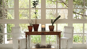 Quarto com uma mesa de madeira, duas cadeiras brancas e uma janela com vista para vegetação.