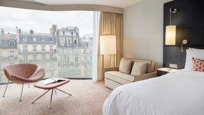 Quarto de hotel com uma janela grande, um sofá, uma poltrona, uma cama e uma mesa de cabeceira com abajur.