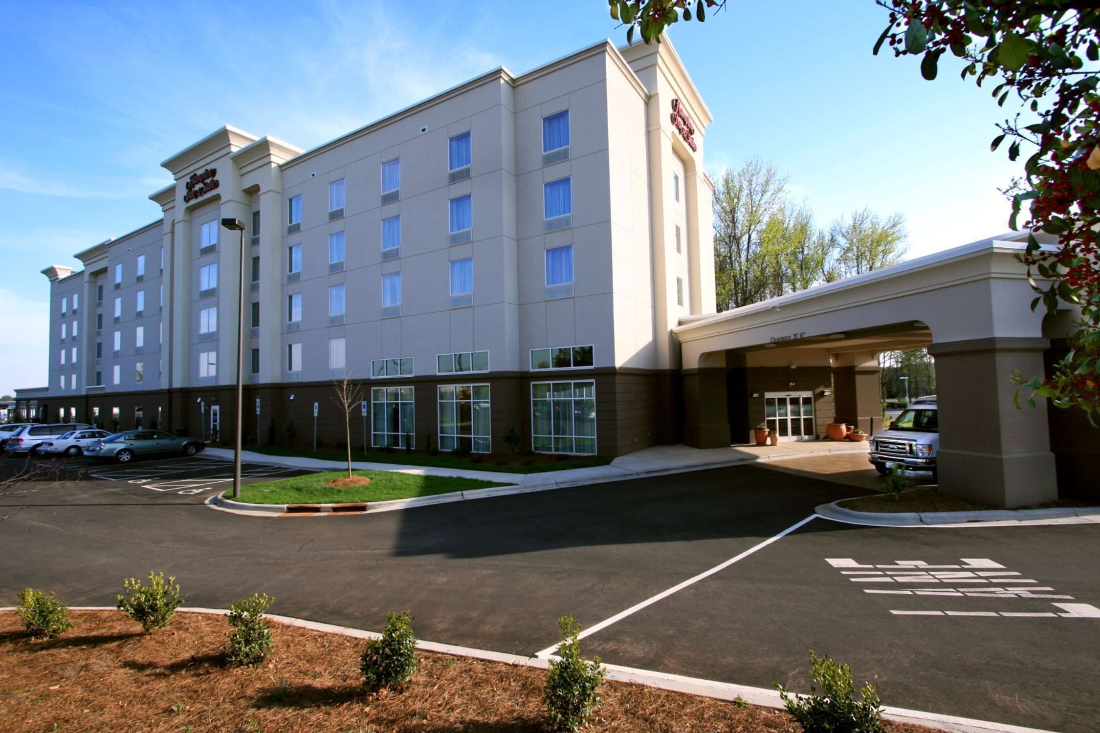 Hotel de vários andares com entrada central, estacionamento e área ajardinada com arbustos.