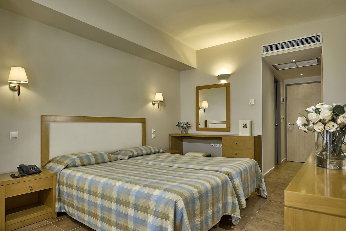 Quarto de hotel com duas camas, mesa de cabeceira com luminária, cômoda, espelho e um vaso com flores.