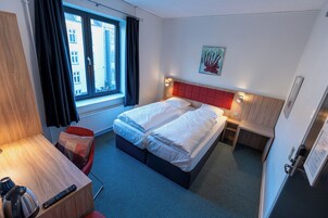 Quarto de hotel com uma cama de casal, uma escrivaninha com chaleira, uma cadeira, uma janela com cortinas e um quadro na parede.