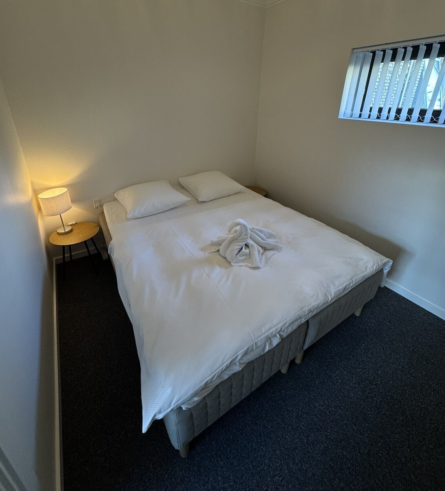Uma cama de solteiro com roupa de cama branca, uma luminária de cabeceira e uma janela com persianas.