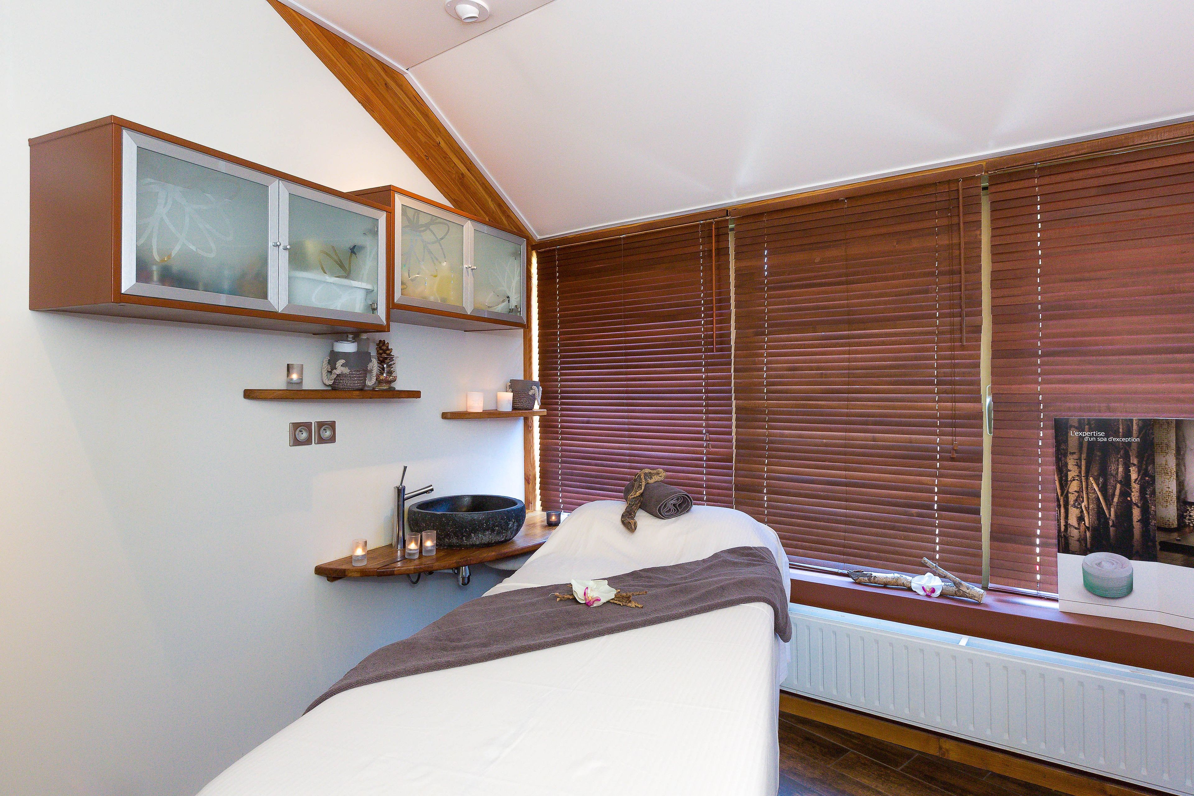Um quarto com cama, janela com persianas, prateleira com uma tigela e armário com portas de vidro.