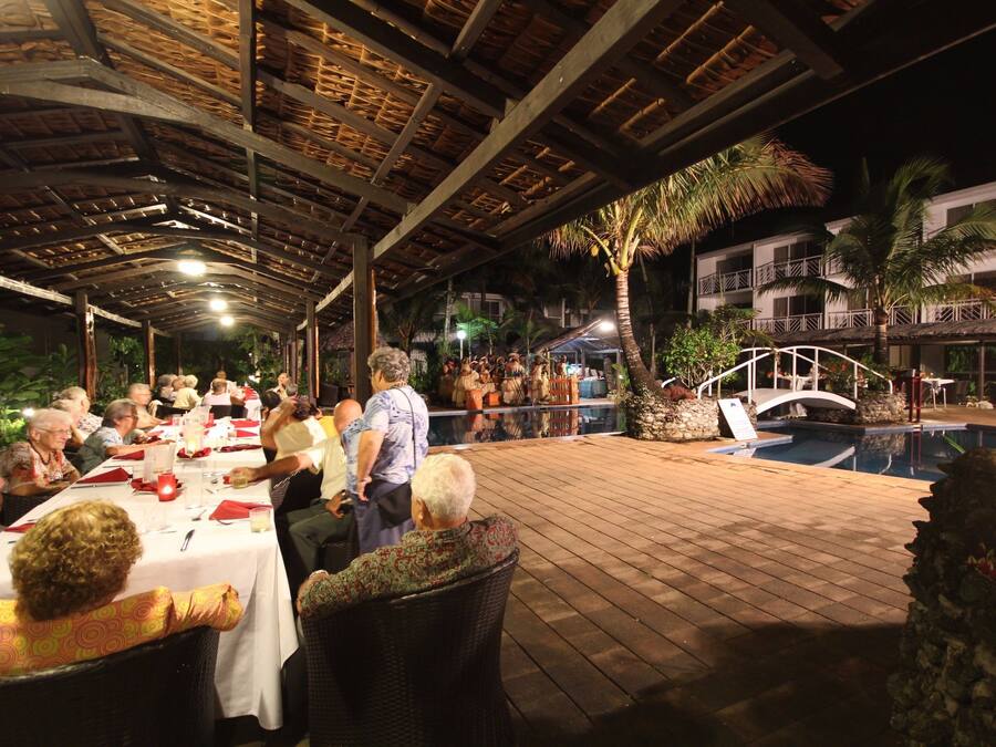 Uma área de refeições ao ar livre com mesas e cadeiras, piscina e um edifício com varandas ao fundo.