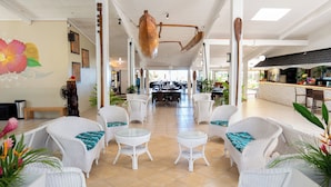 Amplo espaço interno com móveis de vime branco, uma mesa de jantar central e um bar com piso ladrilhado.