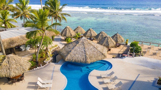 Piscina de resort com formato peculiar, rodeada por espreguiçadeiras e guarda-sóis de palha, com vista para uma praia e oceano.
