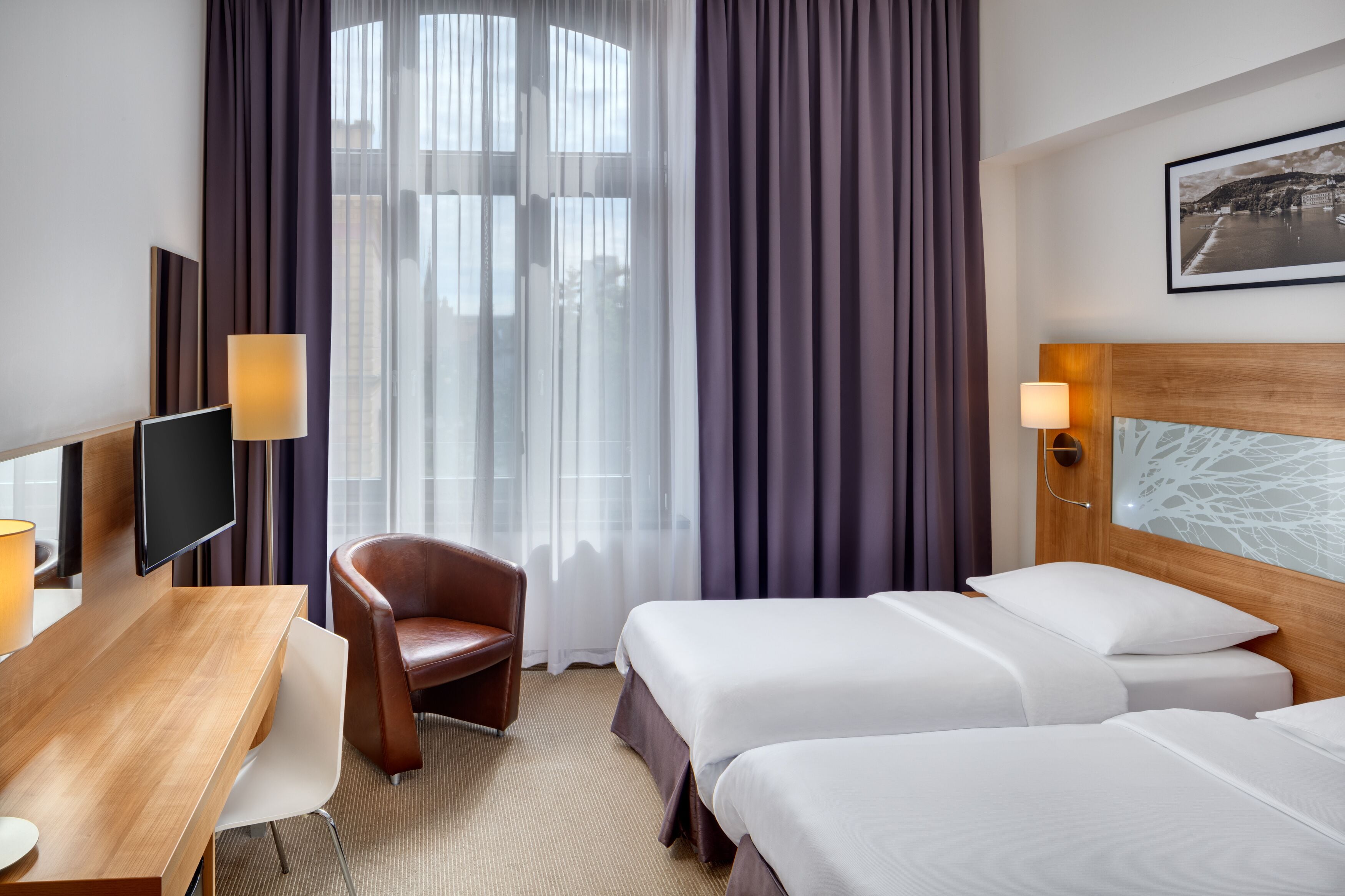 Quarto de hotel com duas camas, uma mesa de trabalho com televisão, uma cadeira e uma janela grande com cortinas.