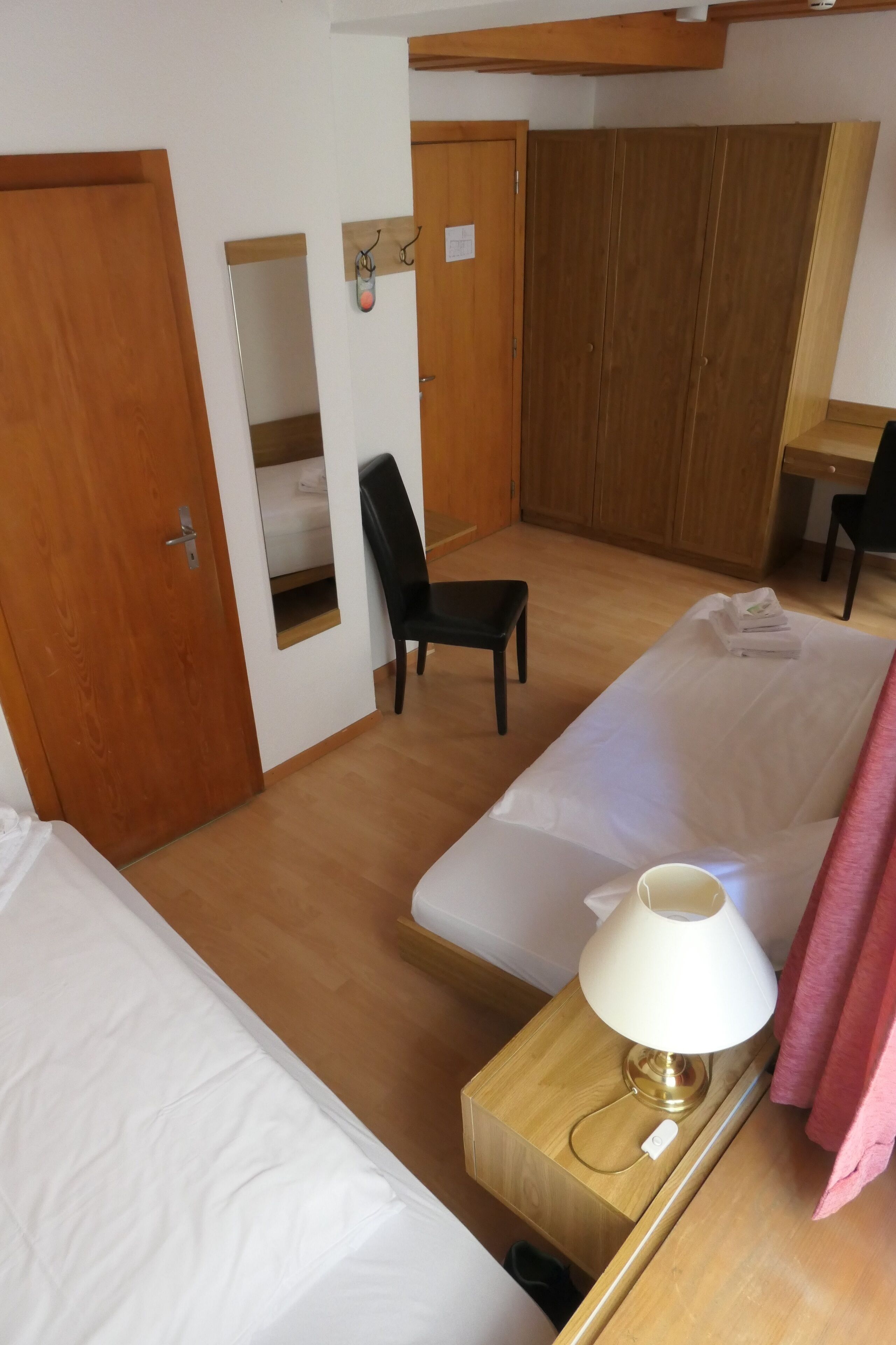 Quarto de hotel com piso de madeira, uma cama com colchão branco, uma cadeira, uma escrivaninha com abajur e um guarda-roupa.