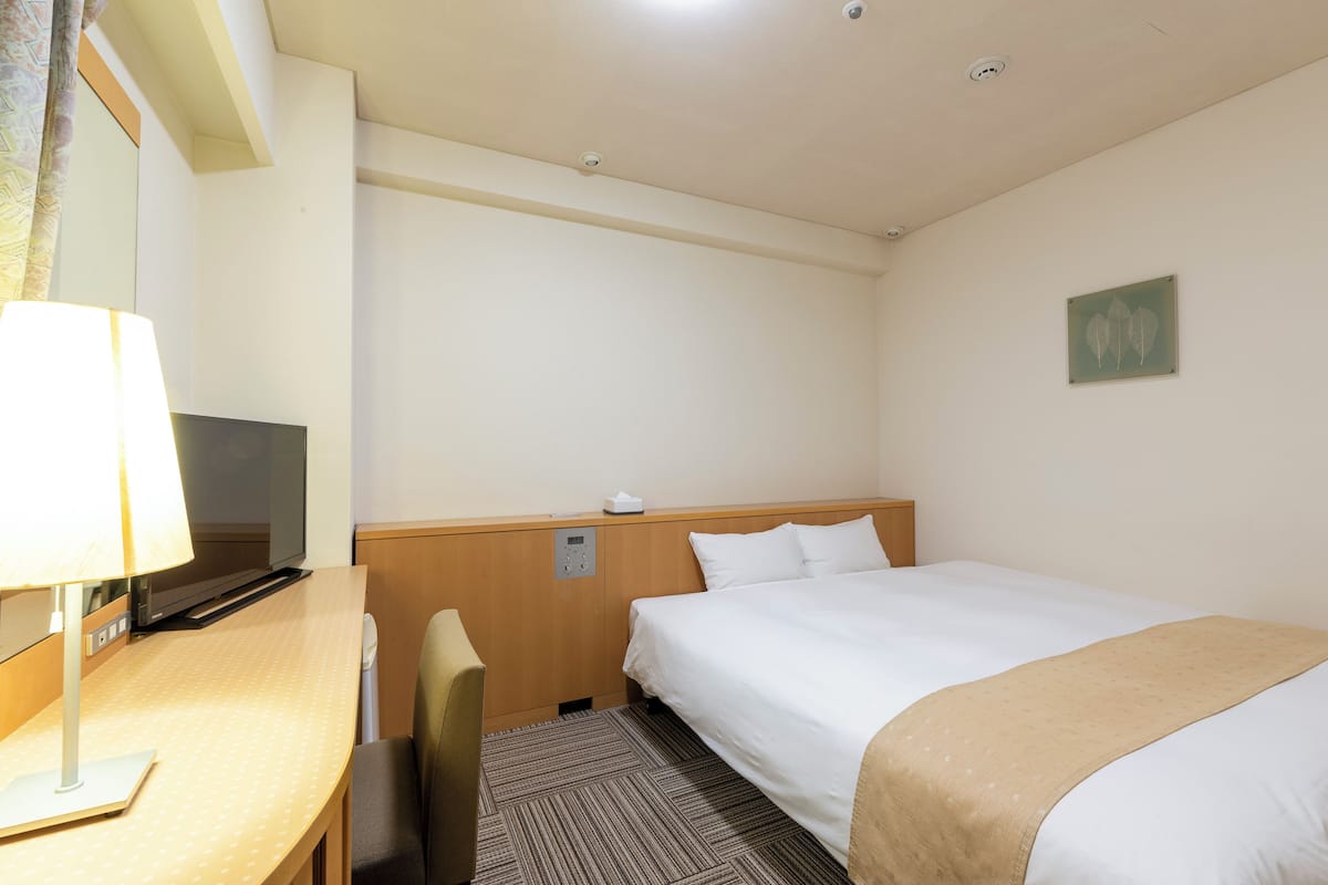 Quarto de hotel com cama, escrivaninha com abajur, televisão e painel de controle fixado na parede.