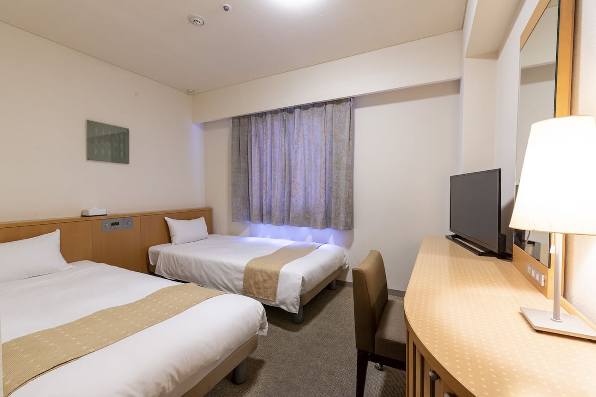 Quarto de hotel com duas camas, uma escrivaninha, uma cadeira, uma televisão e uma janela com cortinas.