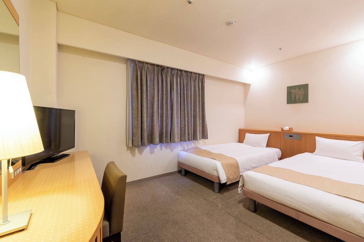 Quarto de hotel com duas camas, uma escrivaninha, televisão e janela com cortinas.