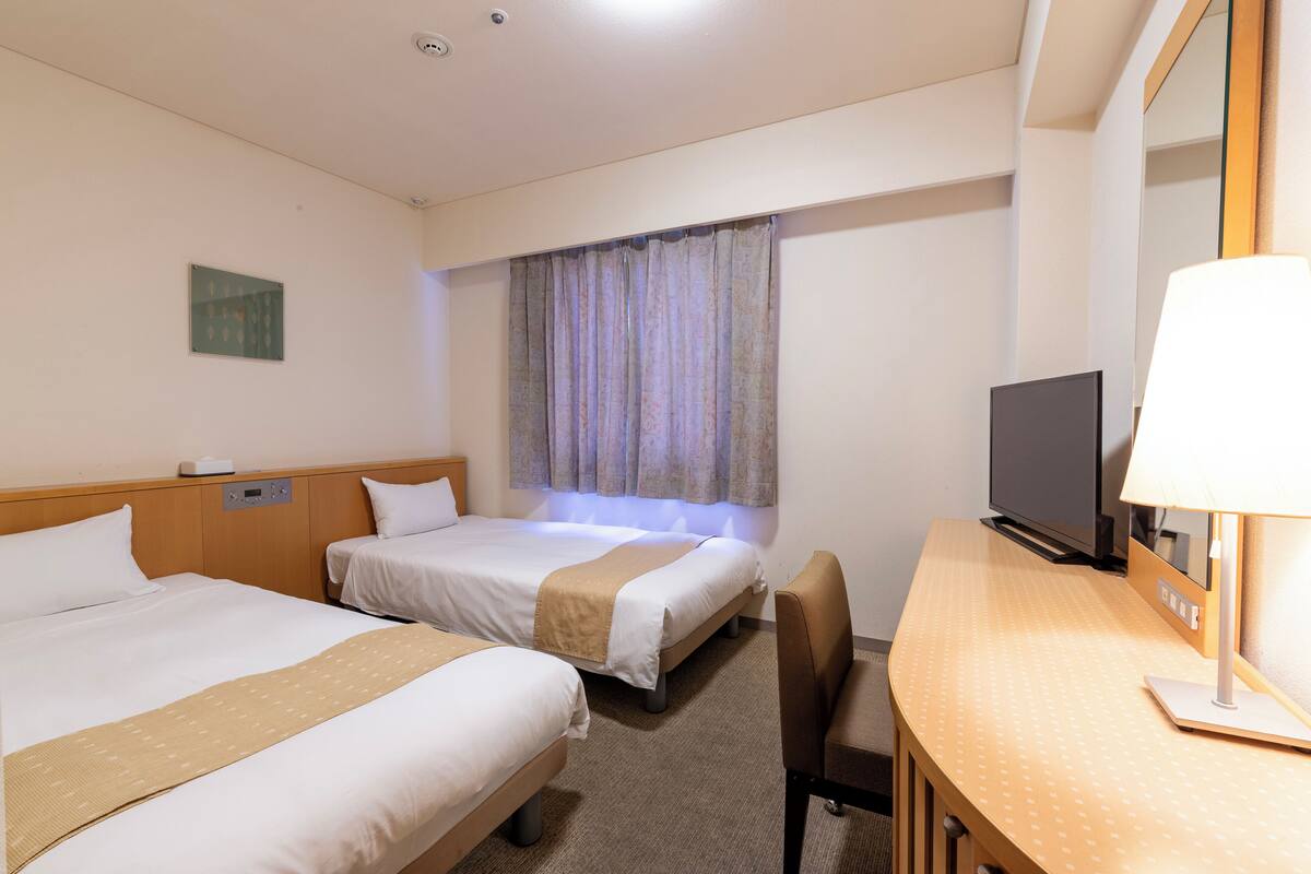 Quarto de hotel com duas camas, uma escrivaninha com abajur, televisão e janela com cortinas.