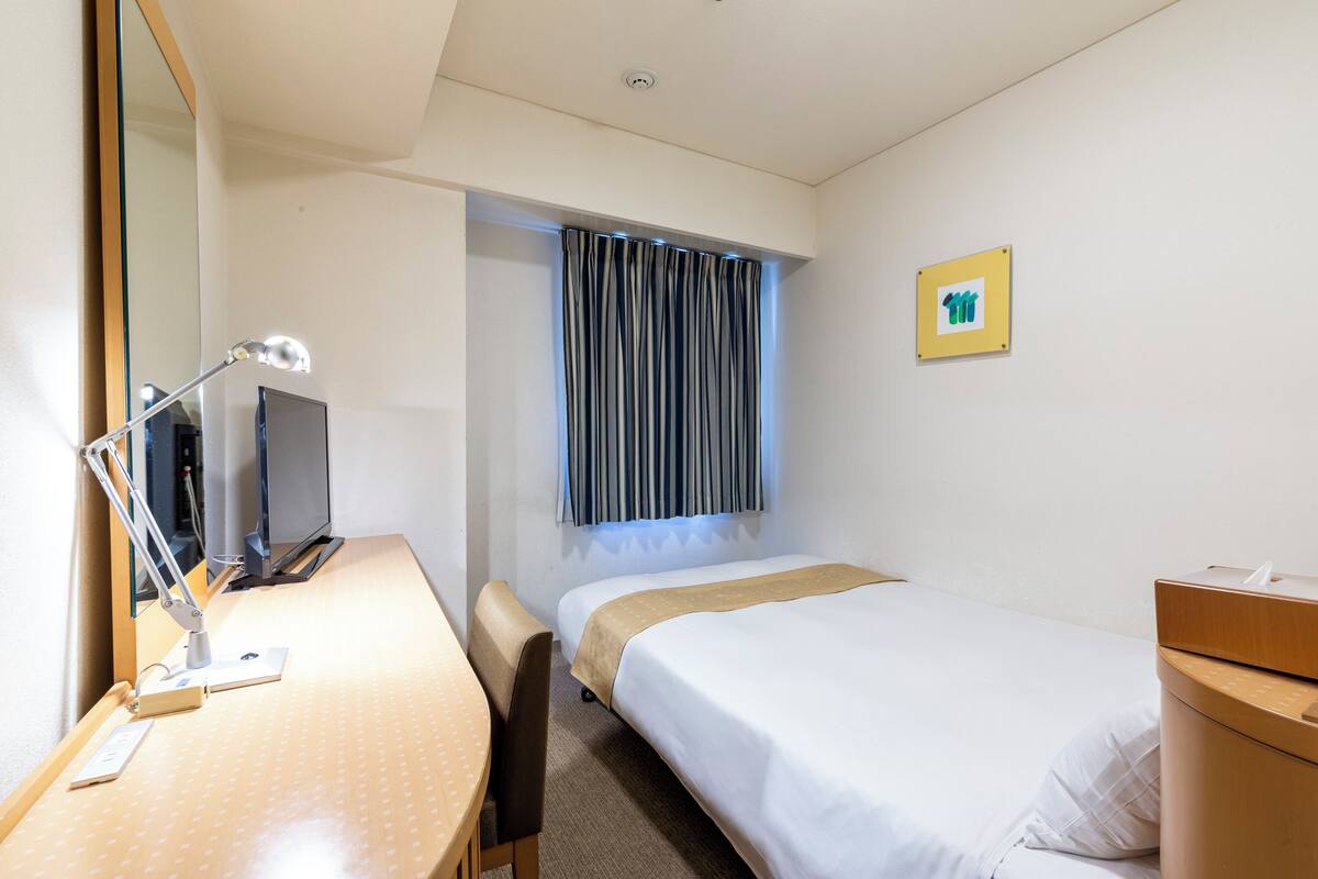 Quarto de hotel com escrivaninha, cama, mesa de cabeceira, espelho, janela com cortinas e um aviso na parede.