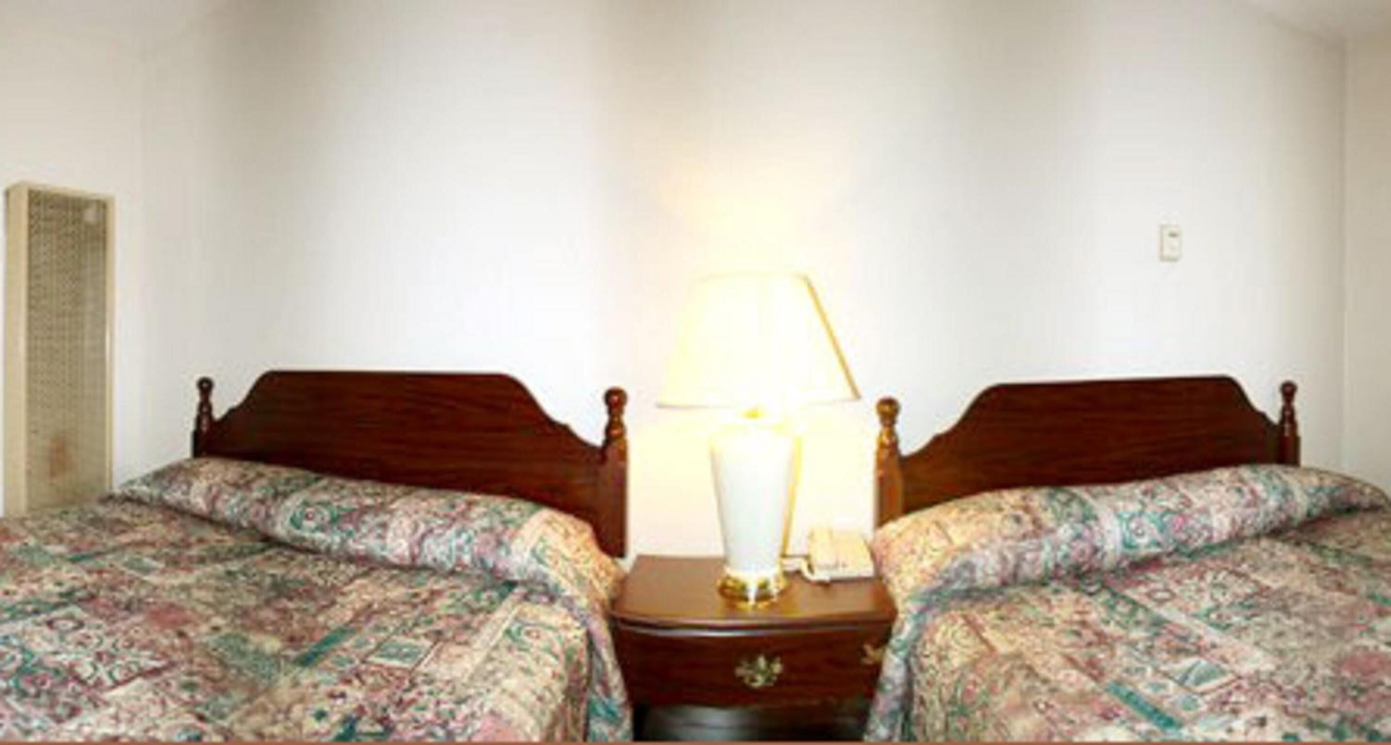 Quarto com duas camas de madeira, cada uma com colcha estampada. Há uma mesa de cabeceira com luminária entre as camas.