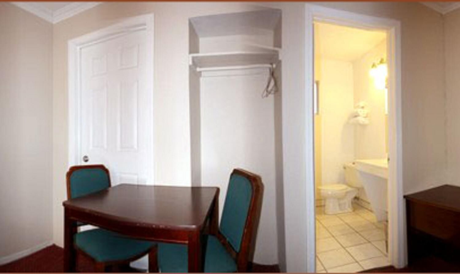 Quarto com mesa de jantar e cadeiras, porta branca e vista para um banheiro com vaso sanitário e pia.