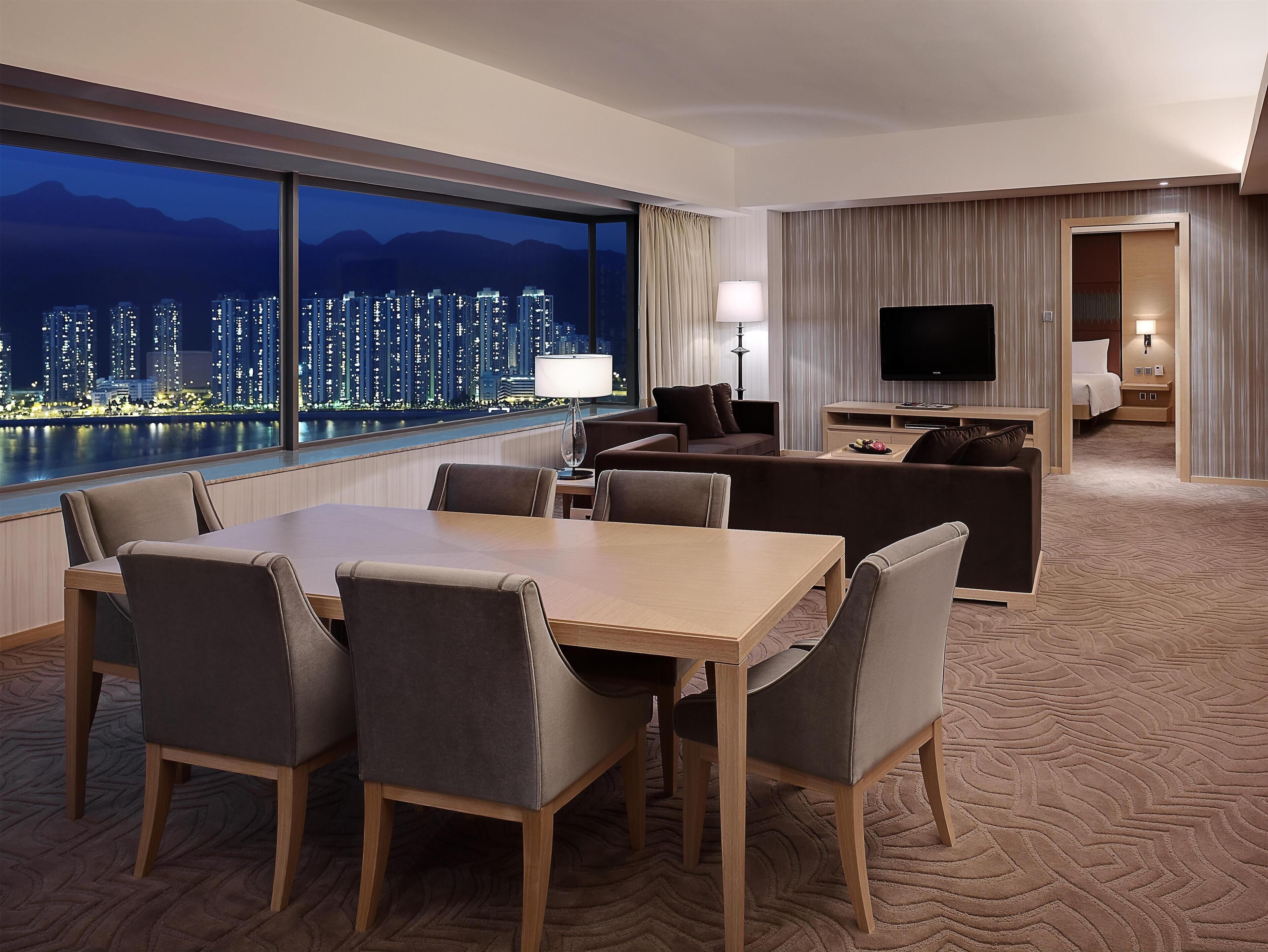 Quarto de hotel com área de jantar, sofá, televisão e vista para a cidade à noite.