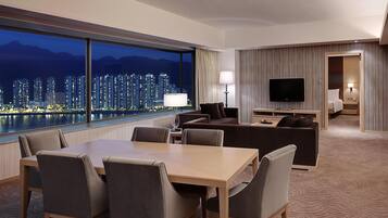 Quarto de hotel com área de jantar, sofá, televisão e vista para a cidade à noite.