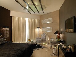 Quarto de hotel moderno com uma cama grande, uma escrivaninha com abajur, uma cadeira com padrão de zebra e vista da cidade por grandes janelas.