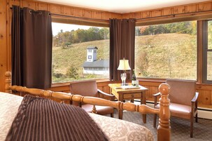 Quarto de hotel com uma cama grande, duas cadeiras de madeira, uma mesa pequena com abajur e vista para uma colina com um edifício.