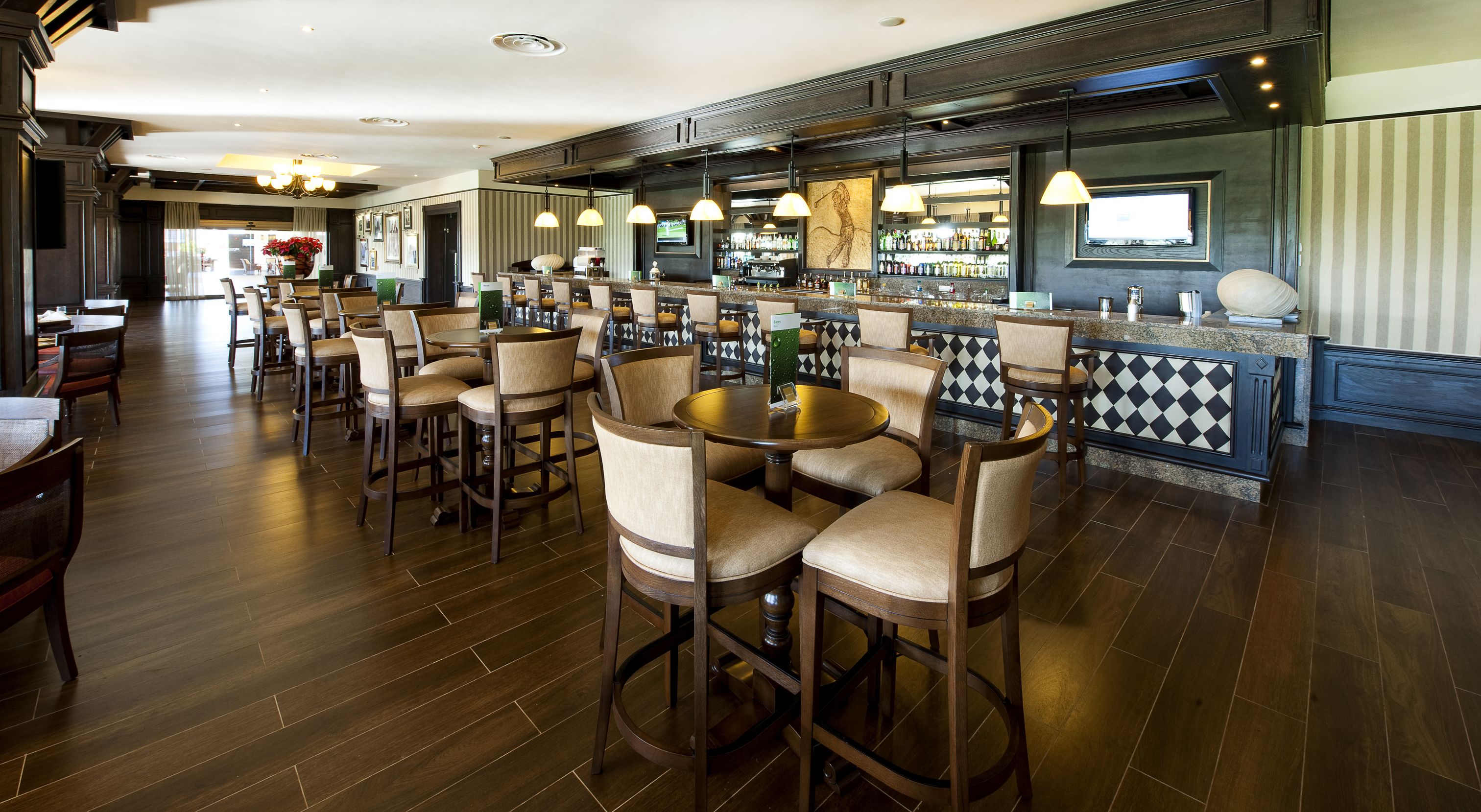 Uma área de bar com mesas redondas e banquetas altas, um balcão com padrão xadrez e uma televisão na parede.