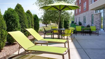 Uma área externa com assentos verdes, cadeiras e uma mesa sob um guarda-sol verde, cercada por árvores e um edifício com grandes janelas.