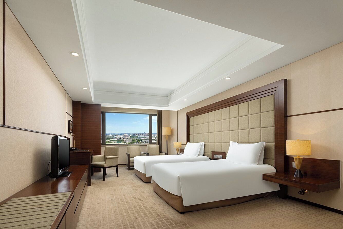 Quarto de hotel com duas camas, uma televisão, uma secretária e uma janela com vista para a cidade.