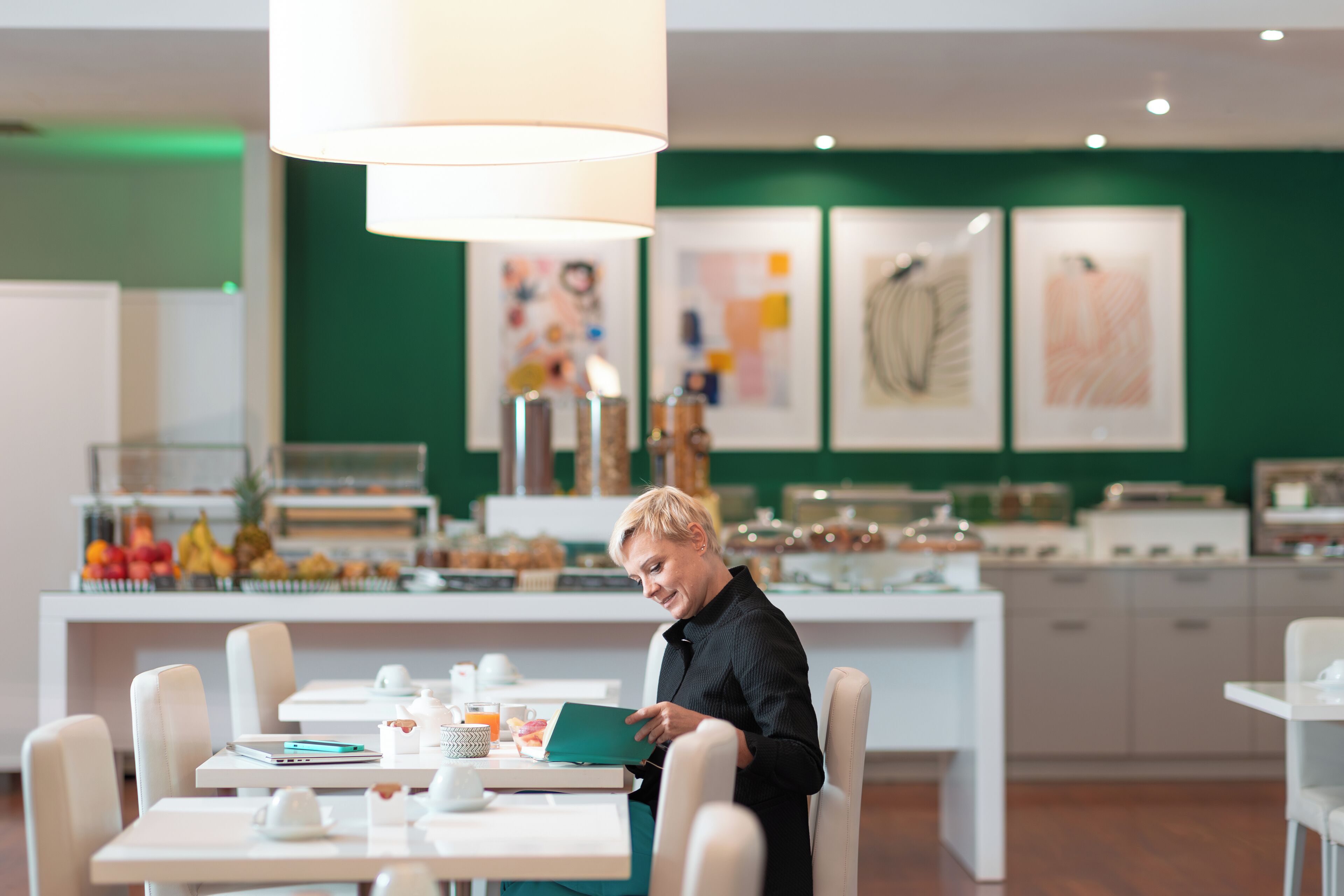 Uma área de refeições moderna com mesas e cadeiras brancas, um balcão central com displays de comida e obras de arte nas paredes.