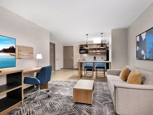 Quarto de hotel moderno com televisão de tela plana, escrivaninha com cadeira, sofá, mesa de centro, tapete estampado, área de cozinha com um banquinho e uma porta que dá para outro cômodo.