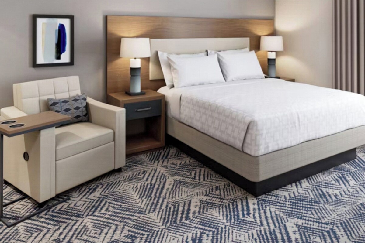 Quarto de hotel com uma cama grande, duas mesas de cabeceira com abajures, uma cadeira, uma escrivaninha pequena e um tapete estampado.