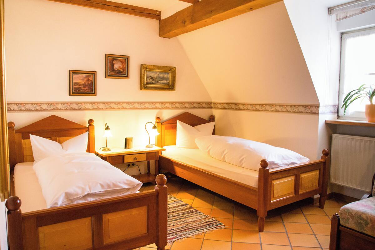 Quarto com duas camas de madeira, uma mesa de cabeceira com abajur e uma janela com planta.