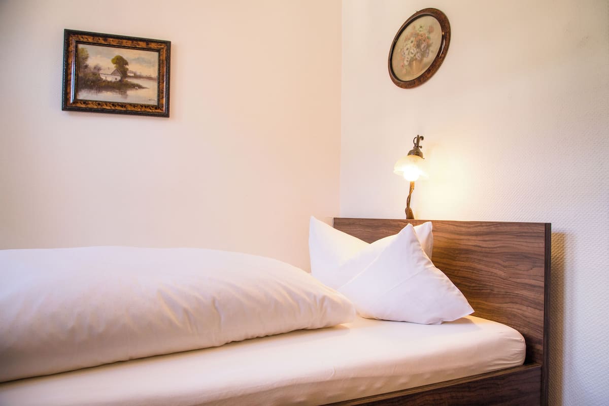 Cama bem arrumada com lençóis e almofadas brancas, cabeceira de madeira, luminária fixada na parede e duas imagens emolduradas.