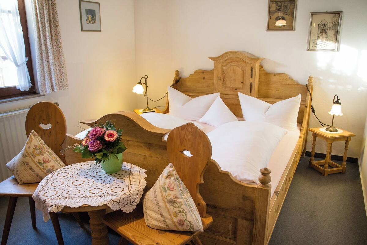 Cama bem arrumada com lençóis brancos, duas mesas de cabeceira com abajures, uma cabeceira de madeira e uma pequena mesa com toalha rendada e um vaso de flores.