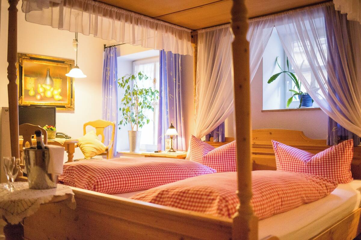 Um quarto acolhedor com cama de madeira, roupa de cama xadrez, mesa de cabeceira com abajur, janela com cortinas e um vaso de planta.