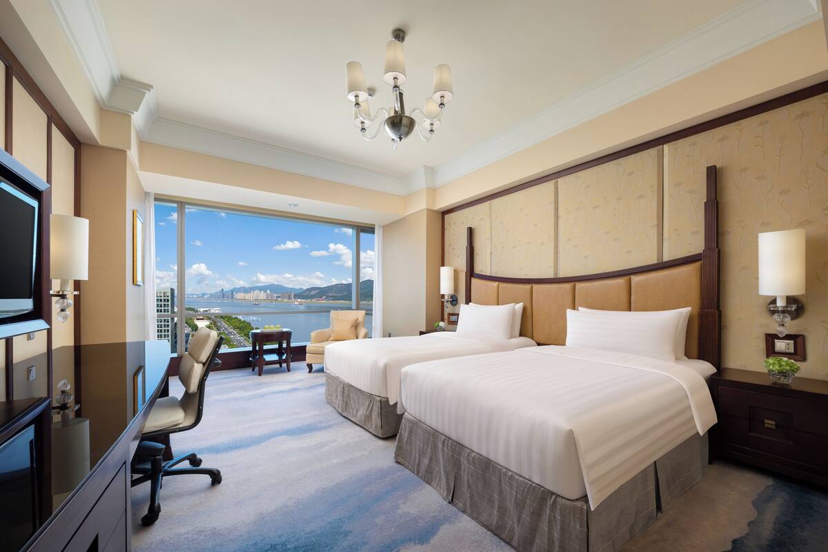 Quarto de hotel com duas camas, uma escrivaninha, televisão e uma janela ampla com vista para a cidade.