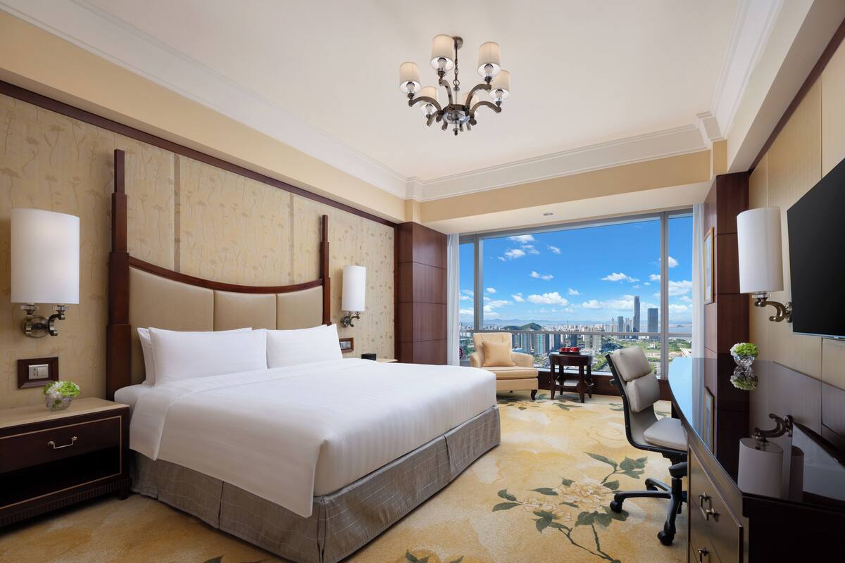 Quarto de hotel espaçoso, com uma cama grande, uma escrivaninha com cadeira, televisão e vista para a cidade.