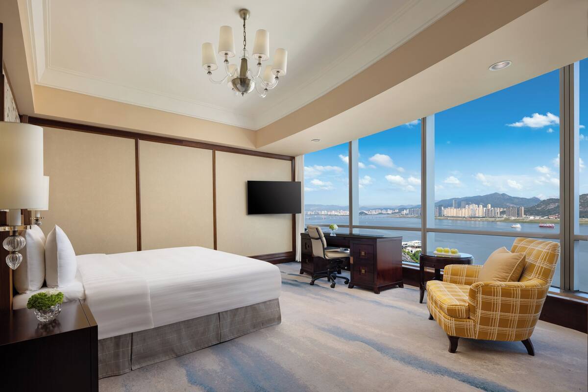 Quarto de hotel com uma cama grande, uma escrivaninha, uma cadeira e televisão na parede.