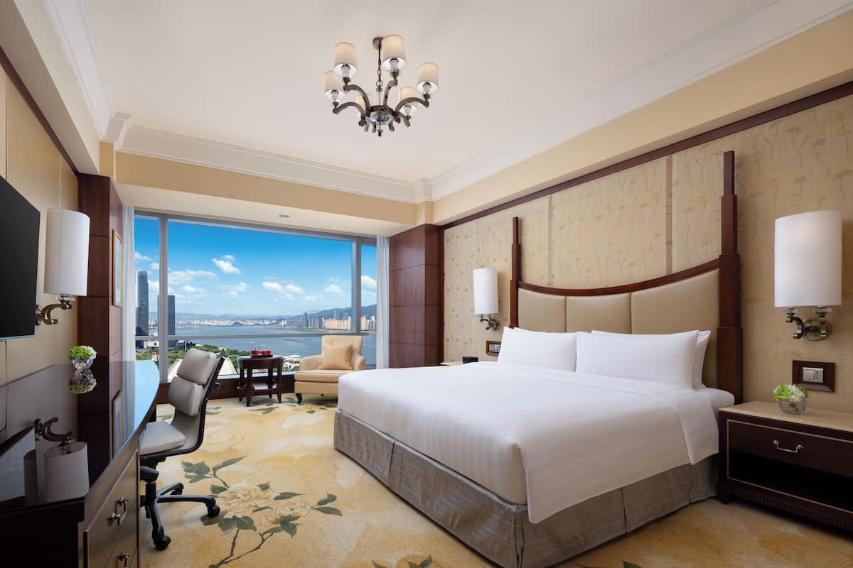Quarto de hotel com uma cama grande, uma escrivaninha com cadeira, televisão e vista para a cidade.