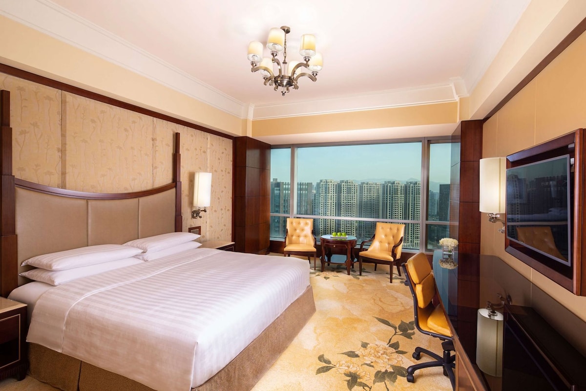 Quarto de hotel com uma cama grande, uma escrivaninha com cadeira, uma mesinha de centro com um vaso, e uma janela ampla com vista para a cidade.