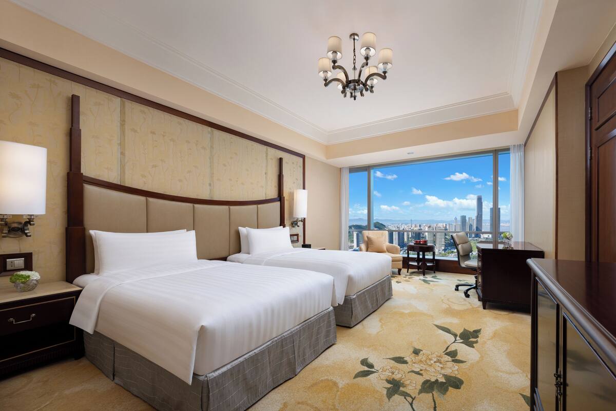 Quarto de hotel com duas camas, uma janela ampla com vista para a cidade, um candelabro e uma mesa de trabalho com uma cadeira.