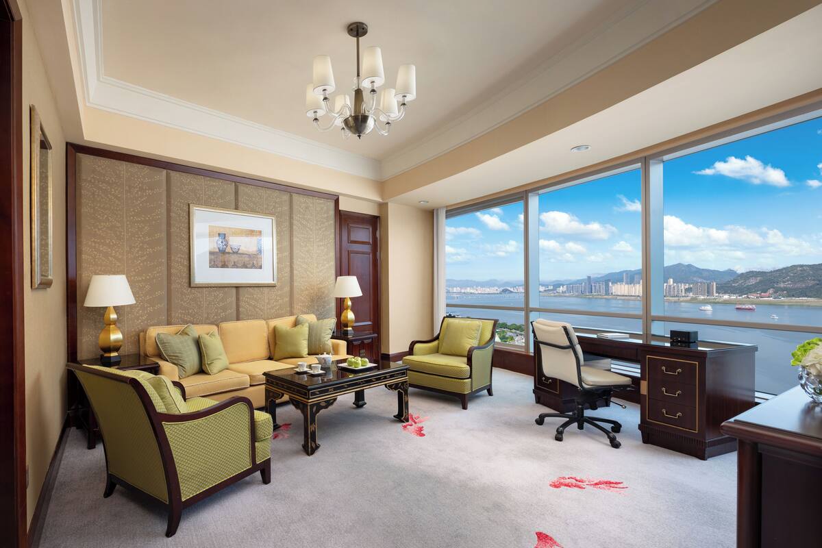 Quarto amplo com uma grande janela que oferece vista para a cidade e para a água. Há um sofá, uma escrivaninha e uma cadeira.