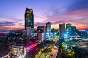 Uma paisagem urbana ao entardecer, com edifícios iluminados e uma rua movimentada.