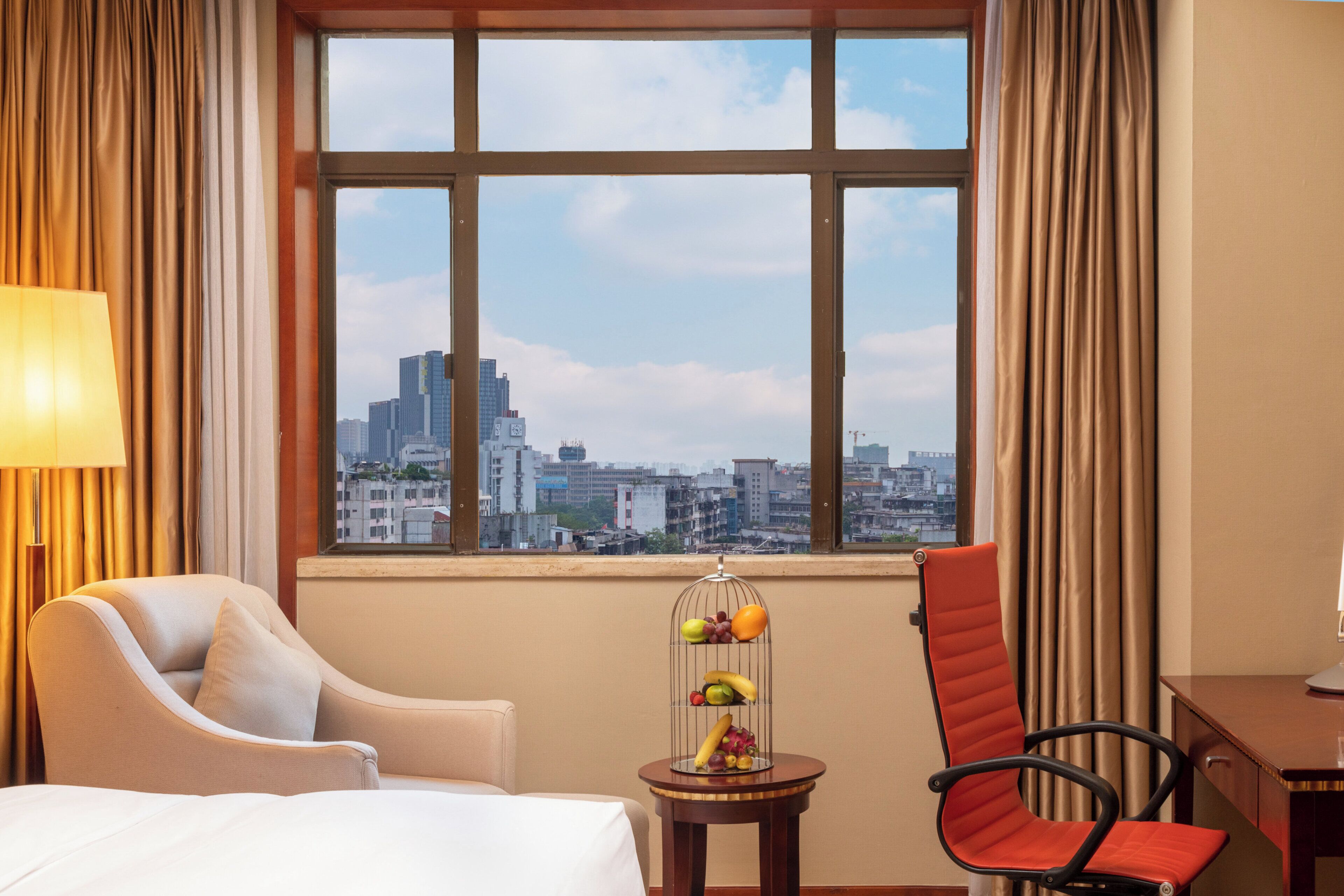 Quarto de hotel com cama, uma cadeira, escrivaninha, abajur, janela com cortinas e uma cesta de frutas.