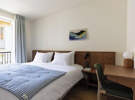Quarto de hotel com cama, uma mesinha, uma cadeira, um quadro na parede e uma janela com cortinas.
