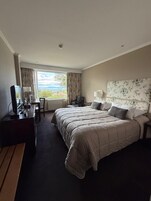 Quarto de hotel com uma cama grande, uma mesa com computador, uma cadeira e uma janela com vista para o exterior.