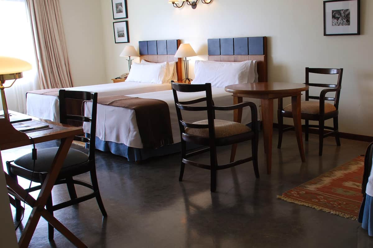 Quarto de hotel com duas camas, uma escrivaninha, uma cadeira, uma mesinha e uma mesa de cabeceira.