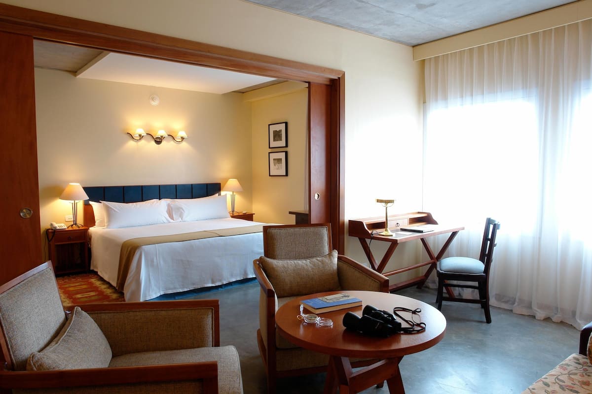 Quarto de hotel com cama, duas cadeiras, uma mesinha, escrivaninha e janela com cortinas.