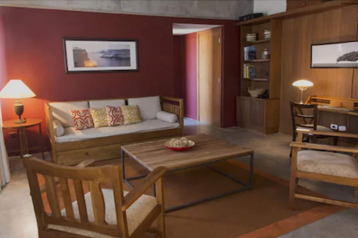 Uma sala de estar aconchegante com um sofá, mesa de centro de madeira e cadeiras. Há uma estante e um abajur em uma mesinha lateral.