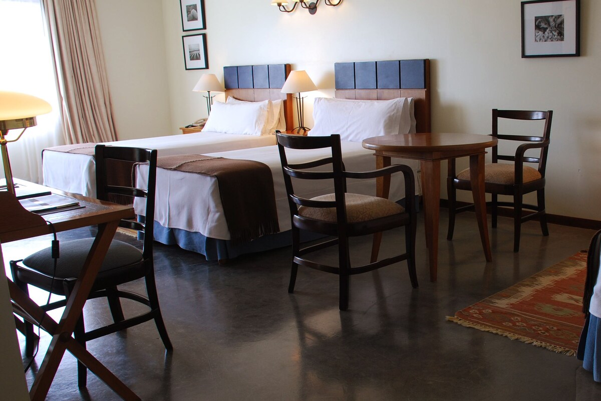 Quarto de hotel com duas camas, uma escrivaninha, duas cadeiras, uma mesinha e uma mesa de cabeceira com luminária.