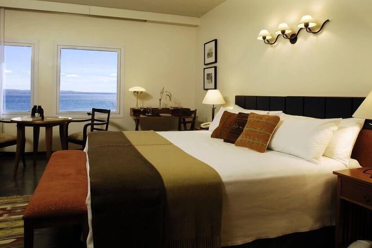 Quarto de hotel com cama, duas cadeiras, escrivaninha, luminária, banco e vista para o mar.