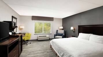 Quarto de hotel com cama, escrivaninha com televisão, cadeira, janela e luminária.